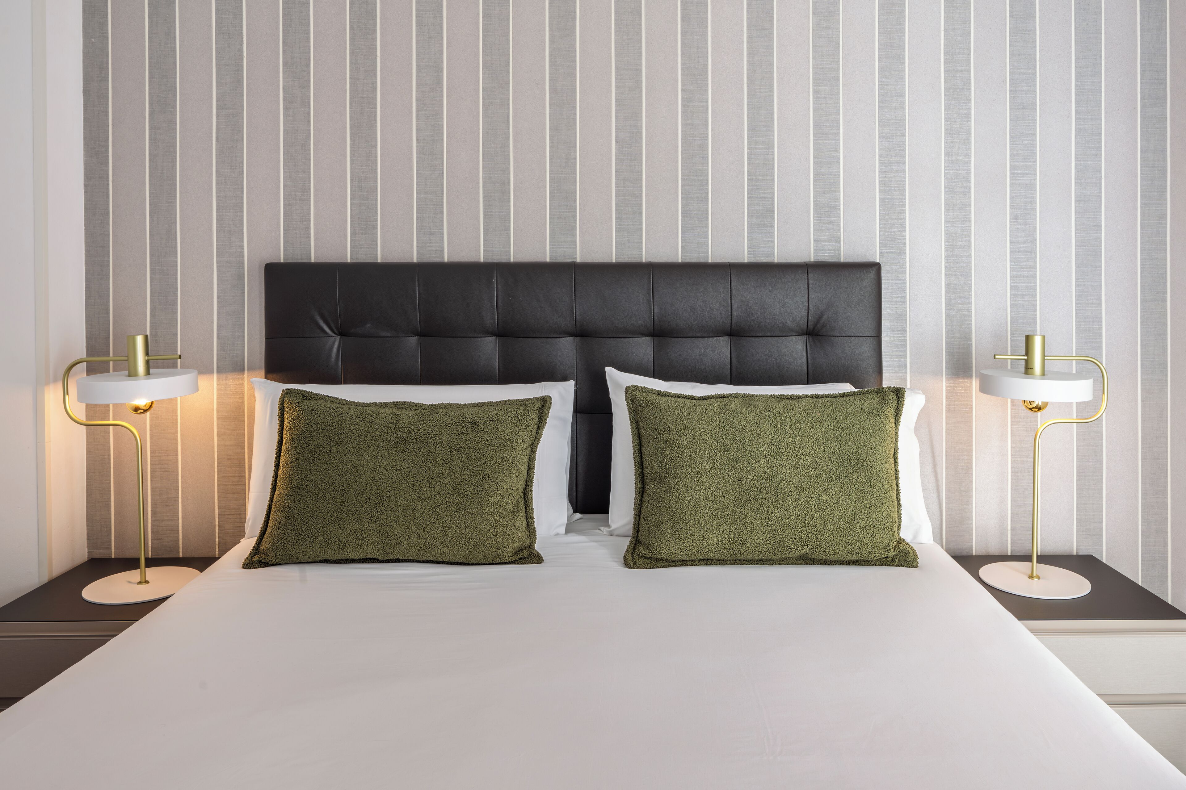 Cama bem arrumada com cabeceira preta, dois travesseiros verdes e duas luminárias de mesa com detalhes dourados.