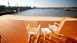 Um deck de madeira com duas cadeiras brancas e uma pequena mesa, com vista para um corpo d'água calmo, com barcos e uma ponte ao fundo.