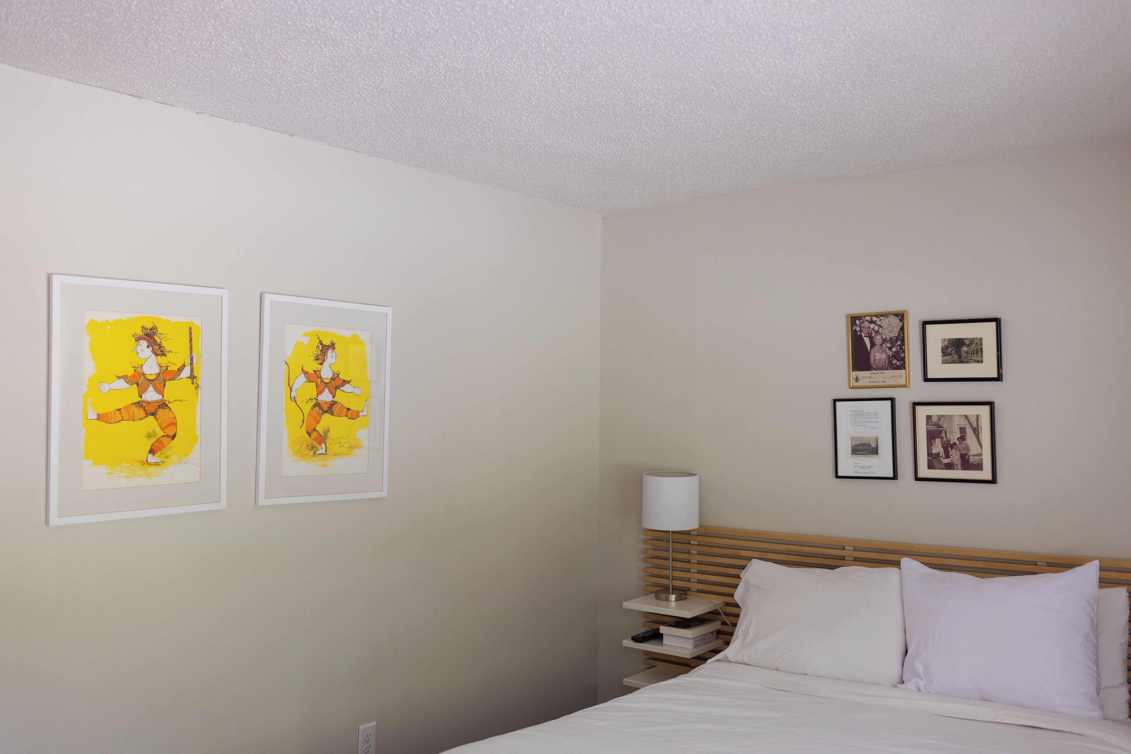 Quarto de hotel com uma cama, mesa de cabeceira, abajur e duas fotos emolduradas na parede.