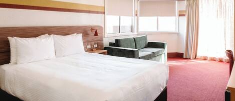 Quarto de hotel com uma cama grande, um sofá verde, uma escrivaninha e uma janela com cortinas.