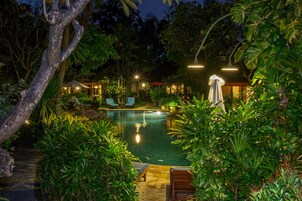 Vista noturna de uma área de piscina rodeada por vegetação exuberante, com espreguiçadeiras e um gazebo.