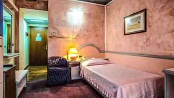 Quarto de hotel com uma cama de solteiro, uma cadeira, uma escrivaninha e um abajur.