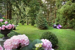 Jardim bem cuidado com diversas árvores, gramado e um vaso grande com flores cor-de-rosa.