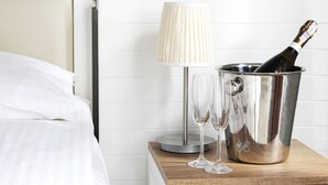 Cama bem arrumada com lençóis brancos, mesa de cabeceira com abajur e balde de champanhe com copos.