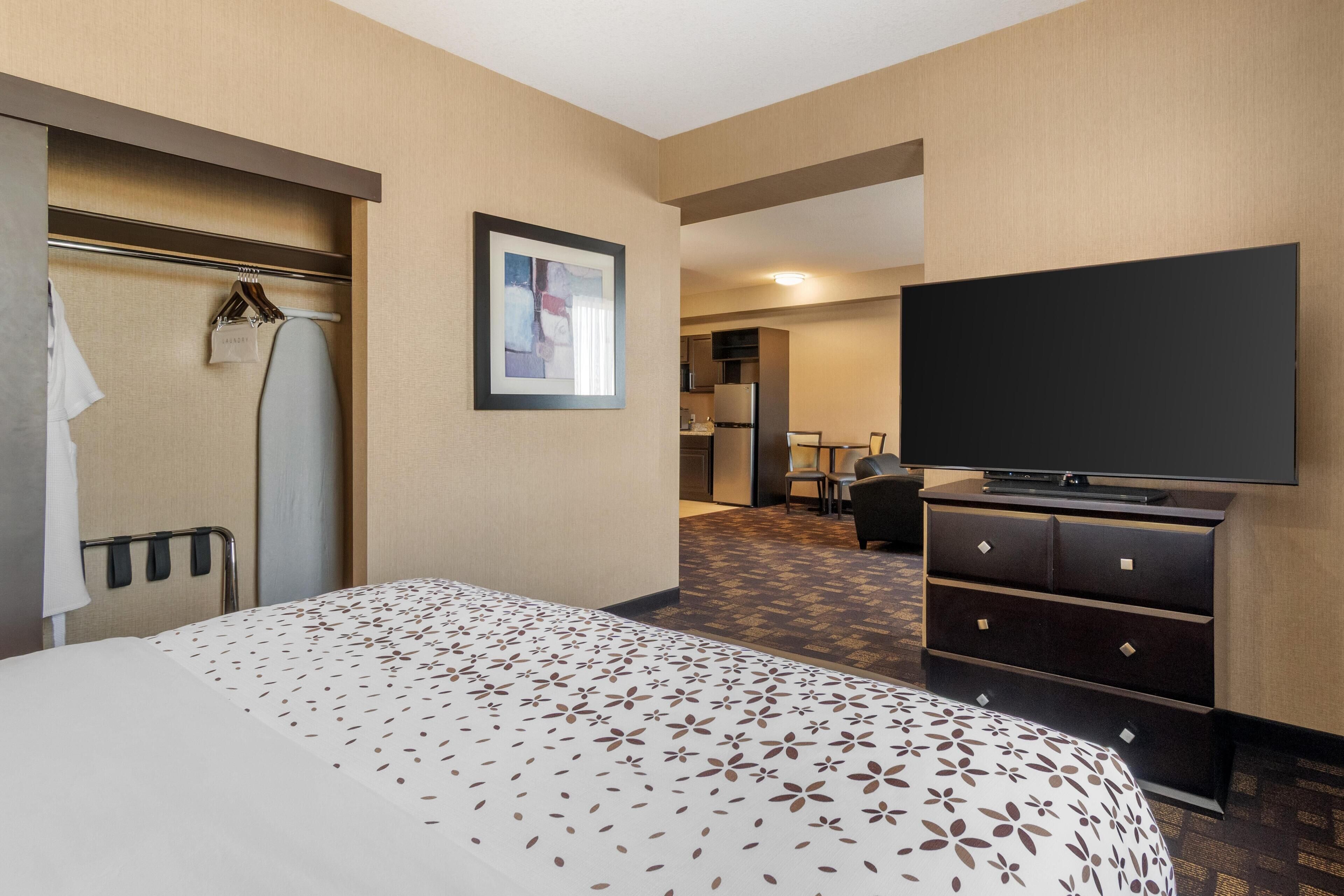 Quarto de hotel com uma cama, televisão, penteadeira, armário e vista para a cozinha.