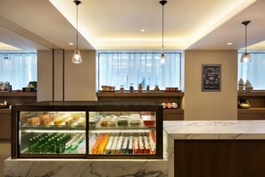 Um hall de hotel com uma recepção de mármore, um refrigerador com bebidas, iluminação pendente e vista da cidade através de grandes janelas.