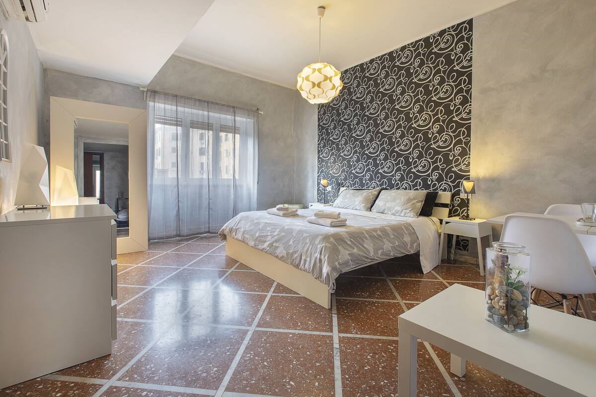 Quarto moderno com uma cama grande, uma escrivaninha e uma cadeira. Uma parede com padrão e uma janela com cortinas.