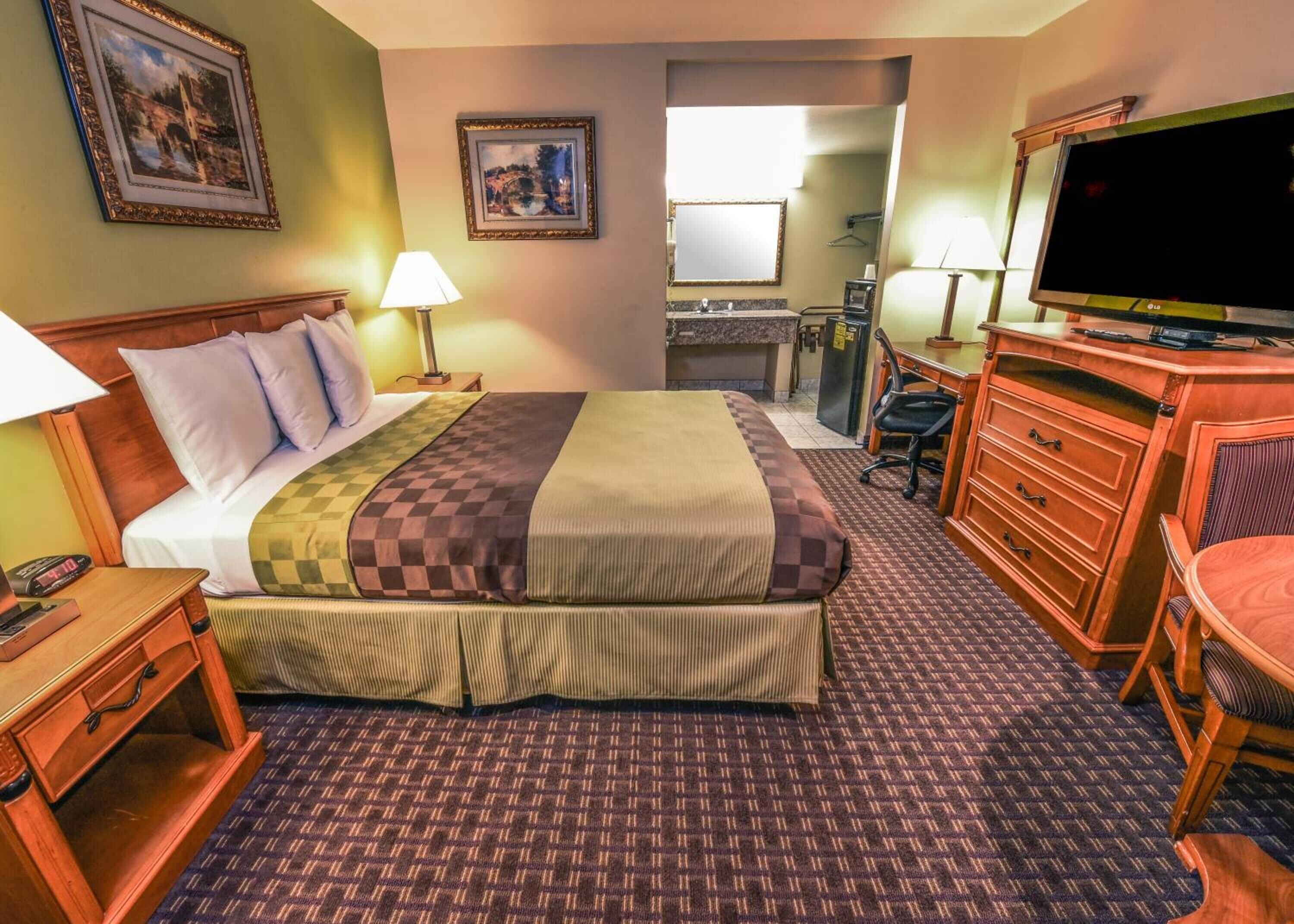 Quarto de hotel com cama, escrivaninha, cadeira, televisão e banheiro visível por uma porta aberta.