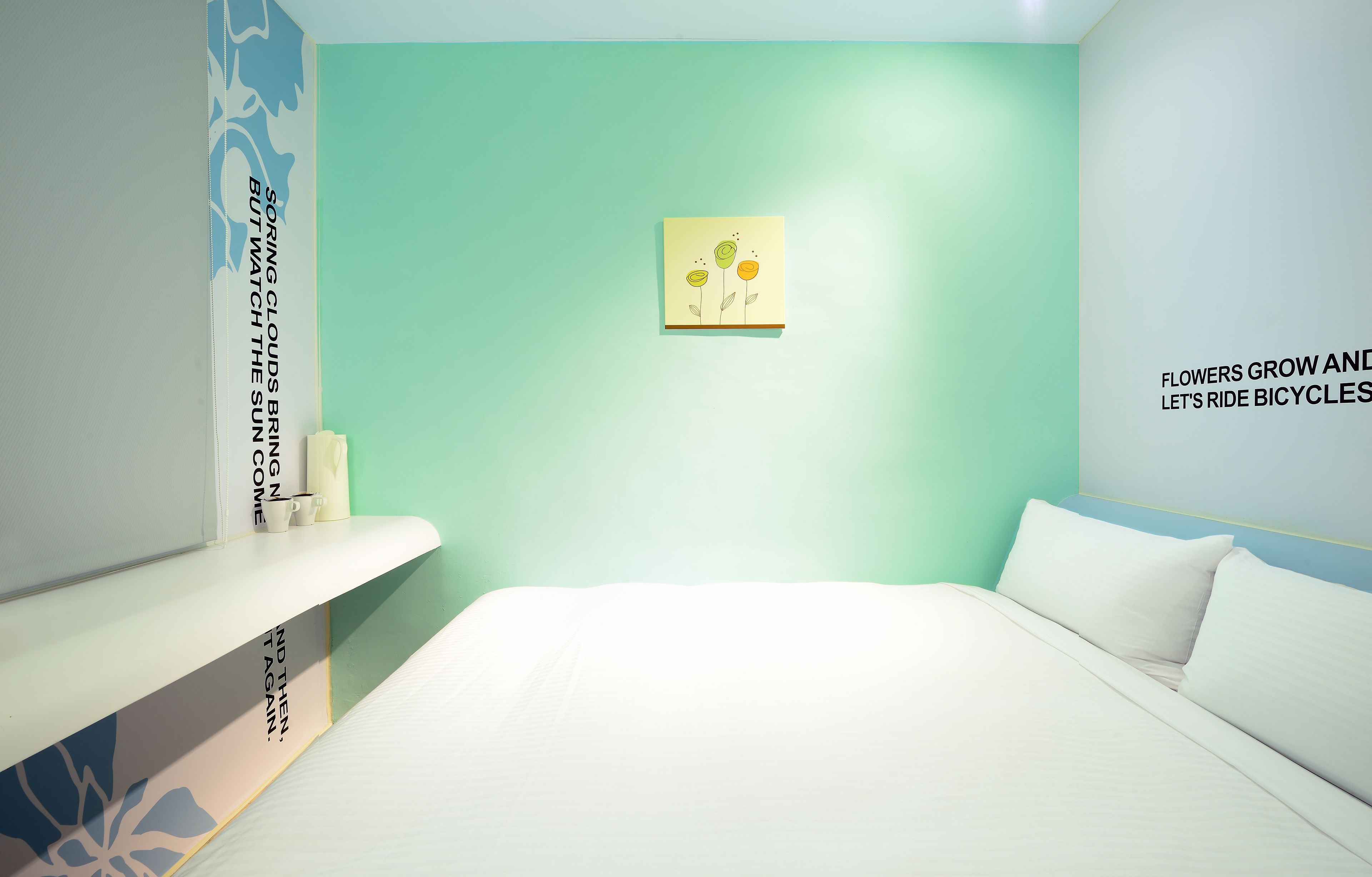 Quarto com cama, mesa de cabeceira e parede decorada com pintura floral e texto motivacional.