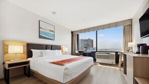 Quarto de hotel com uma cama grande, abajures, uma escrivaninha e vista da cidade através de grandes janelas.