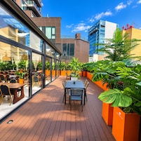 Área externa com varanda, mesas e cadeiras, cercada por vasos de plantas e edifícios modernos.