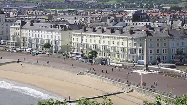 Uma cidade costeira com uma praia, edifícios históricos e vista para as colinas ao fundo.