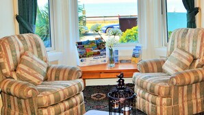 Um quarto com duas poltronas listradas, uma mesa com um vaso e castiçais, e uma janela com vista para o mar e um quadro com 'Lugares para visitar'.