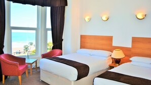 Quarto de hotel com duas camas, uma cadeira vermelha, uma mesinha e uma luminária.