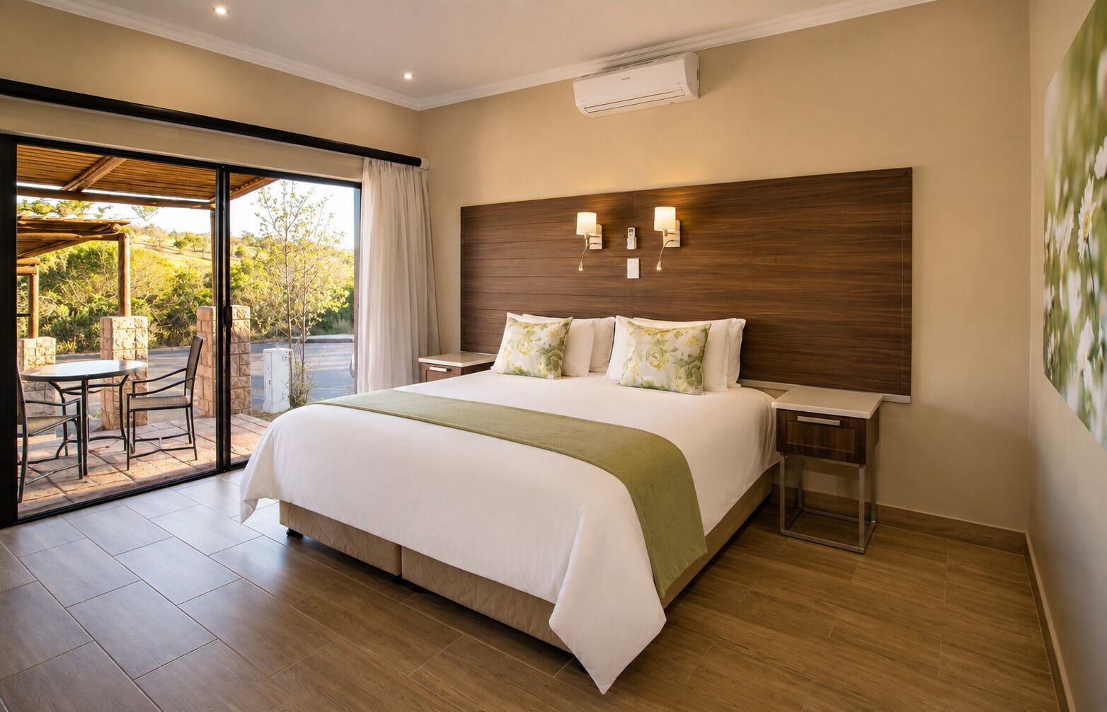 Quarto de hotel com uma cama grande, cabeceira de madeira, porta de vidro que dá para área externa e uma mesinha com cadeiras.