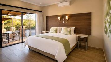 Quarto de hotel com uma cama grande, cabeceira de madeira, porta de vidro que dá para área externa e uma mesinha com cadeiras.