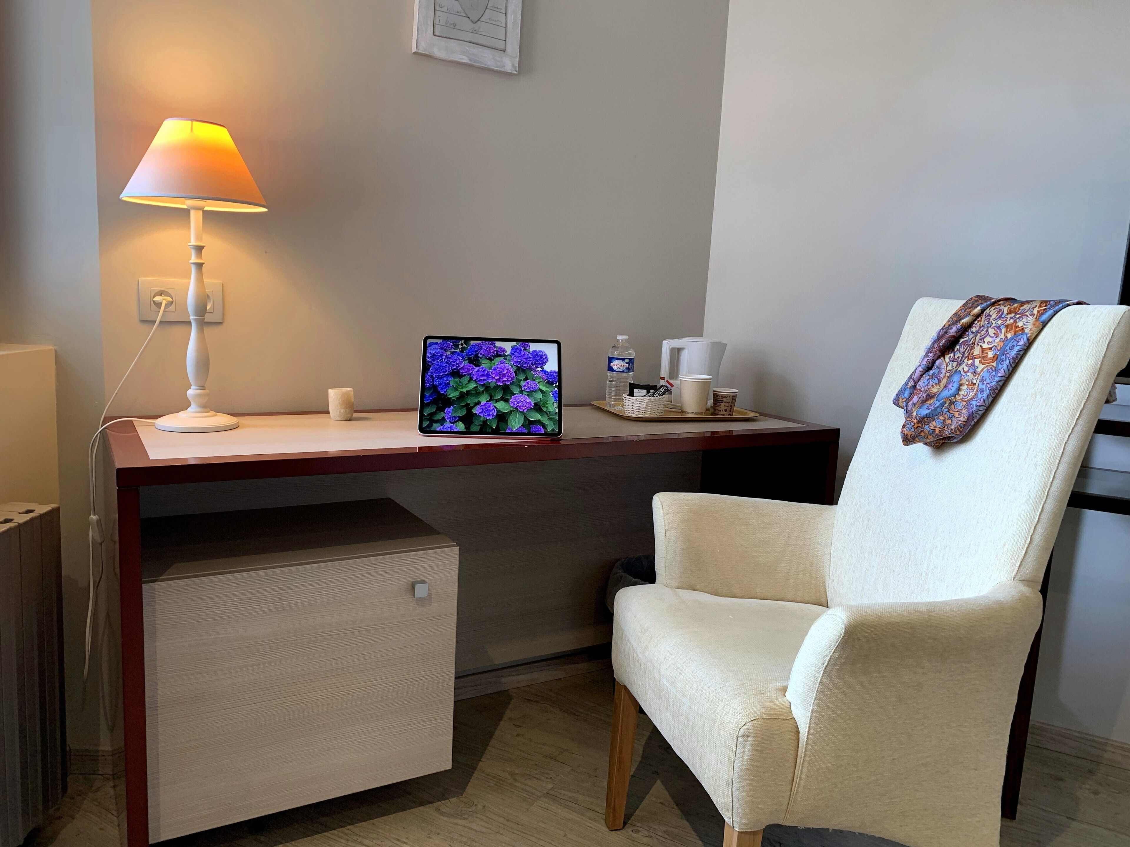 Quarto de hotel com escrivaninha, cadeira, luminária, laptop, e uma mesinha com garrafa de água, xícara de café e caixa de lenços.