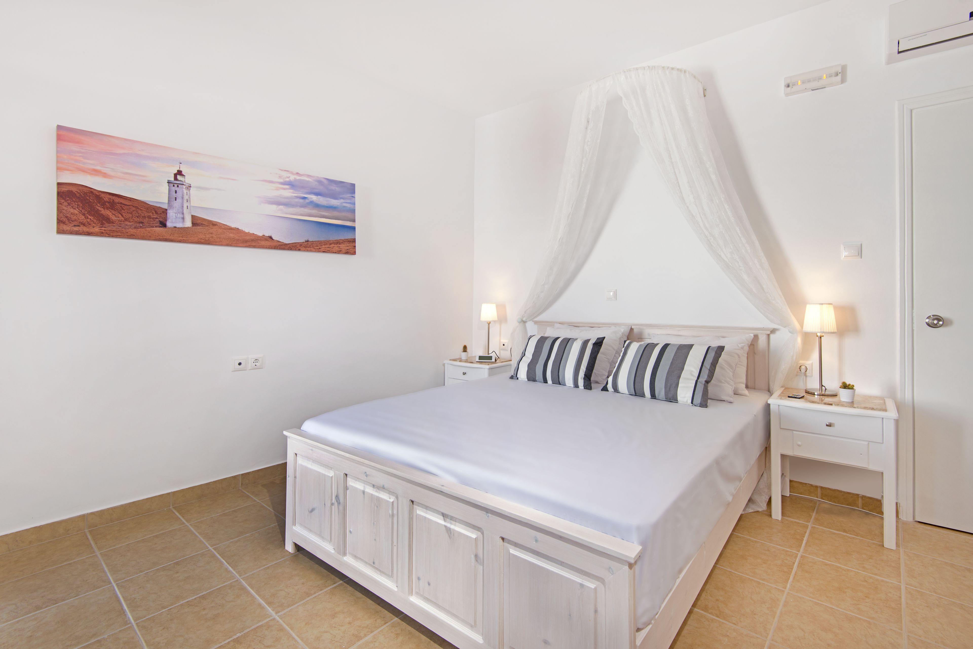 Quarto com cama, mesas de cabeceira, luminária de cabeceira, ar condicionado fixado na parede e uma pintura de um farol.