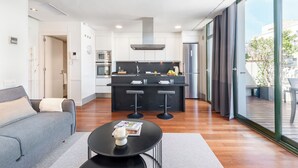 Uma cozinha moderna com ilha central, área de jantar com sofá e mesa de centro, e janelas amplas com vista para o exterior.