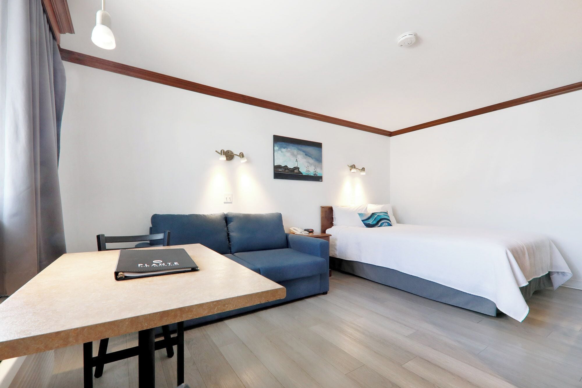 Quarto de hotel com cama, sofá azul, mesa de madeira e duas luzes fixadas na parede.