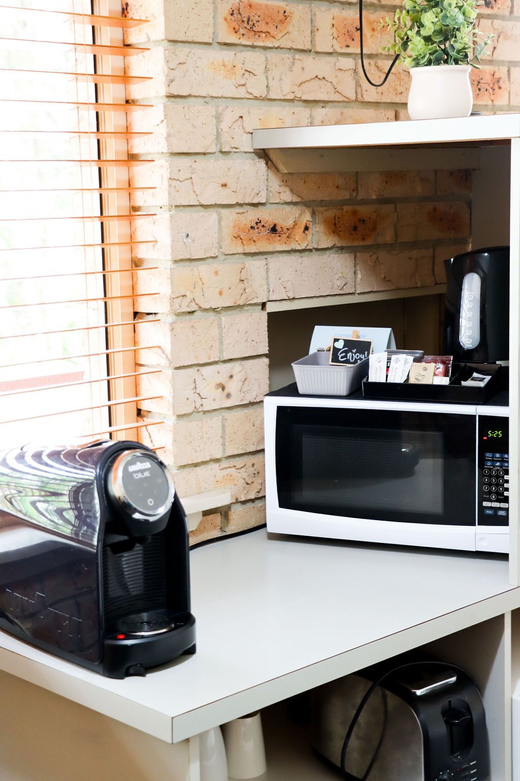 Uma cozinha com cafeteira, micro-ondas e uma pequena prateleira com uma planta e alguns objetos.