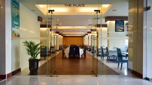 Um saguão de hotel com uma mesa de recepção, uma grande planta em vaso e uma área de refeições com mesas e cadeiras.