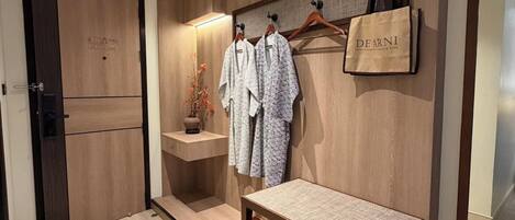 Quarto de hotel com guarda-roupa de madeira, um banco, um porta-toalhas com roupões e uma pequena mesa com um vaso de flores.