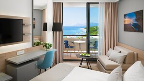Quarto de hotel moderno com uma cama grande, uma escrivaninha, uma cadeira, uma mesinha com maçãs e uma varanda com vista para o mar.