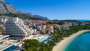 Uma vila costeira com um grande hotel com vários andares, piscina e uma praia de águas azul-claras.