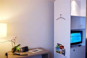 Quarto de hotel com escrivaninha, luminária, televisão, suporte para revistas e cabideiro.