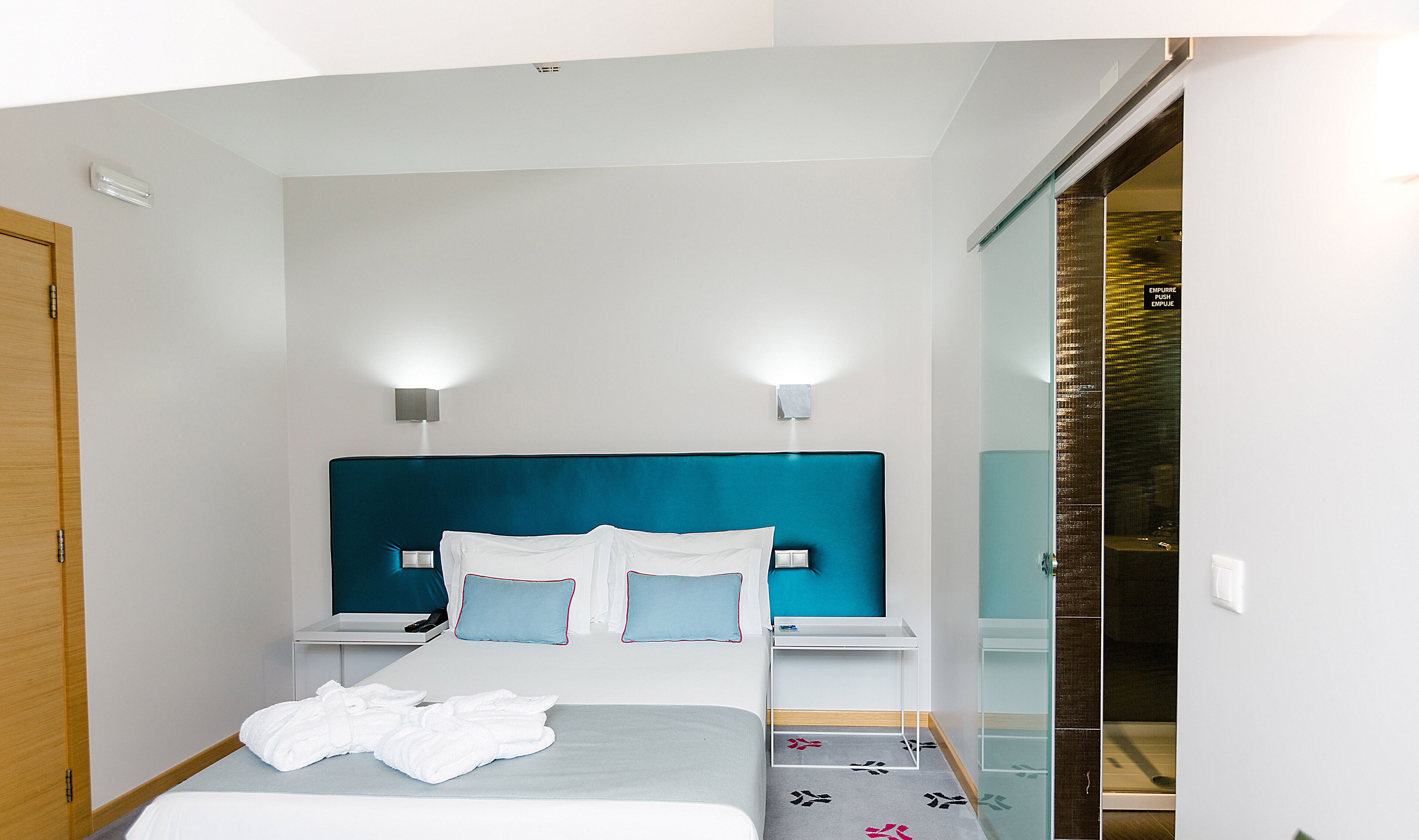 Quarto de hotel moderno com uma cama grande, duas mesas de cabeceira e um painel de parede de vidro.