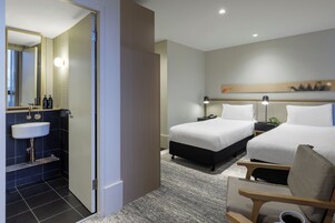 Quarto de hotel com banheiro, duas camas, uma cadeira e uma mesa de cabeceira.
