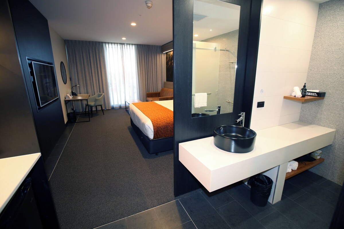 Quarto de hotel moderno com uma cama grande, uma escrivaninha, uma cadeira, banheiro com pia e espelho, e chuveiro.
