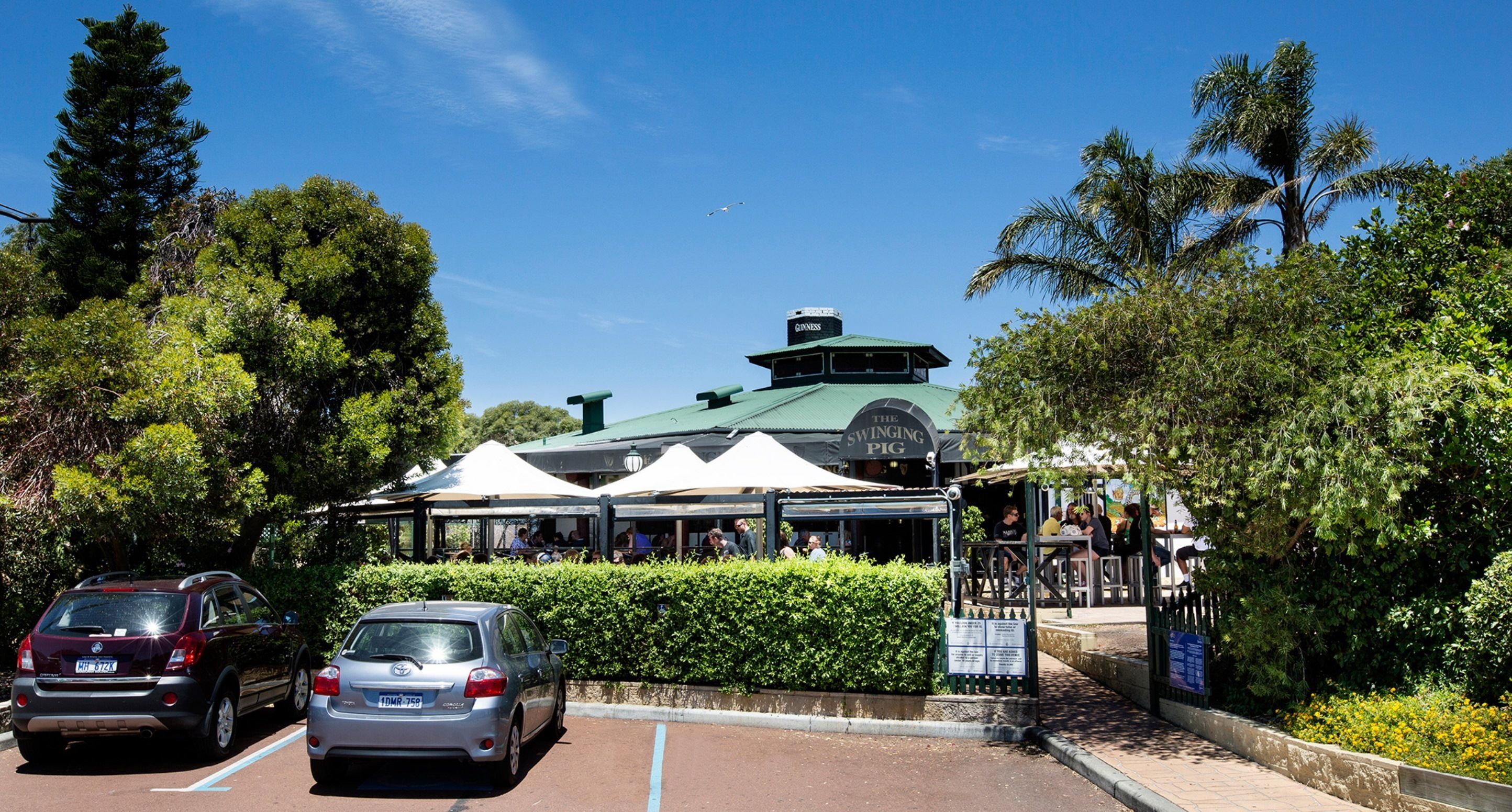 Restaurante com mesas ao ar livre, telhado verde e placa com o texto 'The Swinging Pig'.
