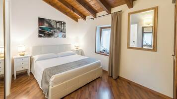 Um quarto com uma cama grande, piso de madeira, teto com vigas aparentes, um quadro na parede, uma janela com cortinas e um espelho.