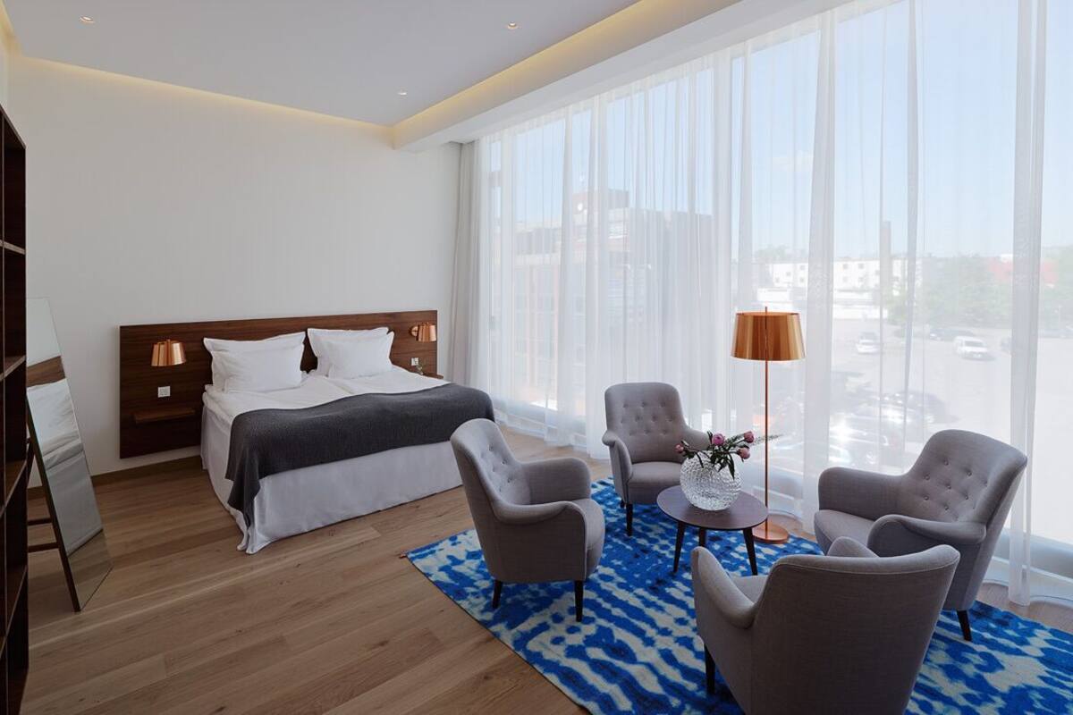 Quarto de hotel moderno com uma cama grande, duas poltronas, uma mesa redonda pequena e vista para o exterior através de cortinas leves.