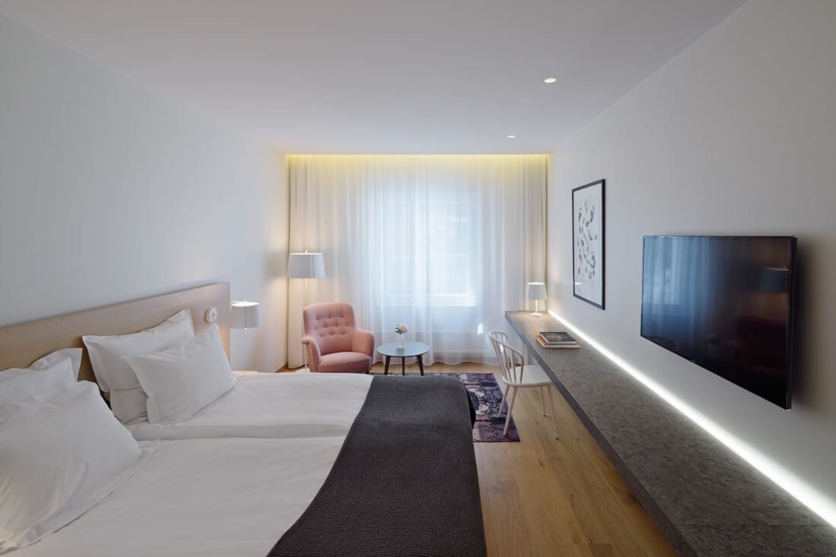 Quarto de hotel com cama, escrivaninha, cadeira, televisão e janela com cortinas.
