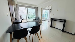 Uma sala de estar moderna com uma mesa de jantar preta, duas cadeiras pretas, um poltrona cinzenta e uma janela grande com vista para edifícios.