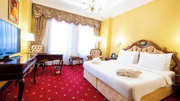 Quarto de hotel com cama grande, escrivaninha, duas cadeiras, televisão e um candelabro.