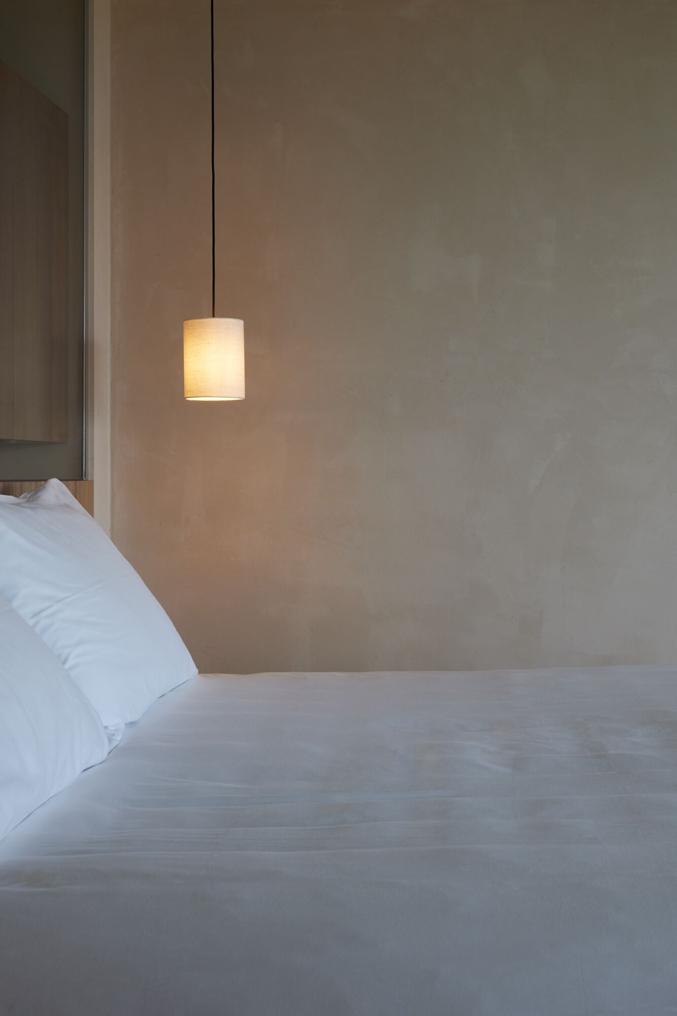 Cama bem arrumada com lençóis brancos em quarto com parede lisa e luminária pendente.