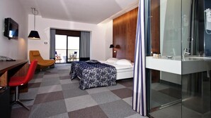 Quarto de hotel moderno com chão xadrez, cama com colcha estampada, escrivaninha com cadeira, televisão e chuveiro de vidro.