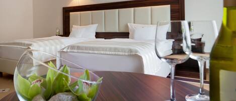 Quarto de hotel com uma cama grande, duas luminárias de cabeceira, um painel de cabeceira em madeira e uma mesa com uma tigela de suculentas e dois copos de vinho.