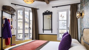 Quarto de hotel com cama, janela com cortinas, um cabideiro com casaco e um espelho na parede.
