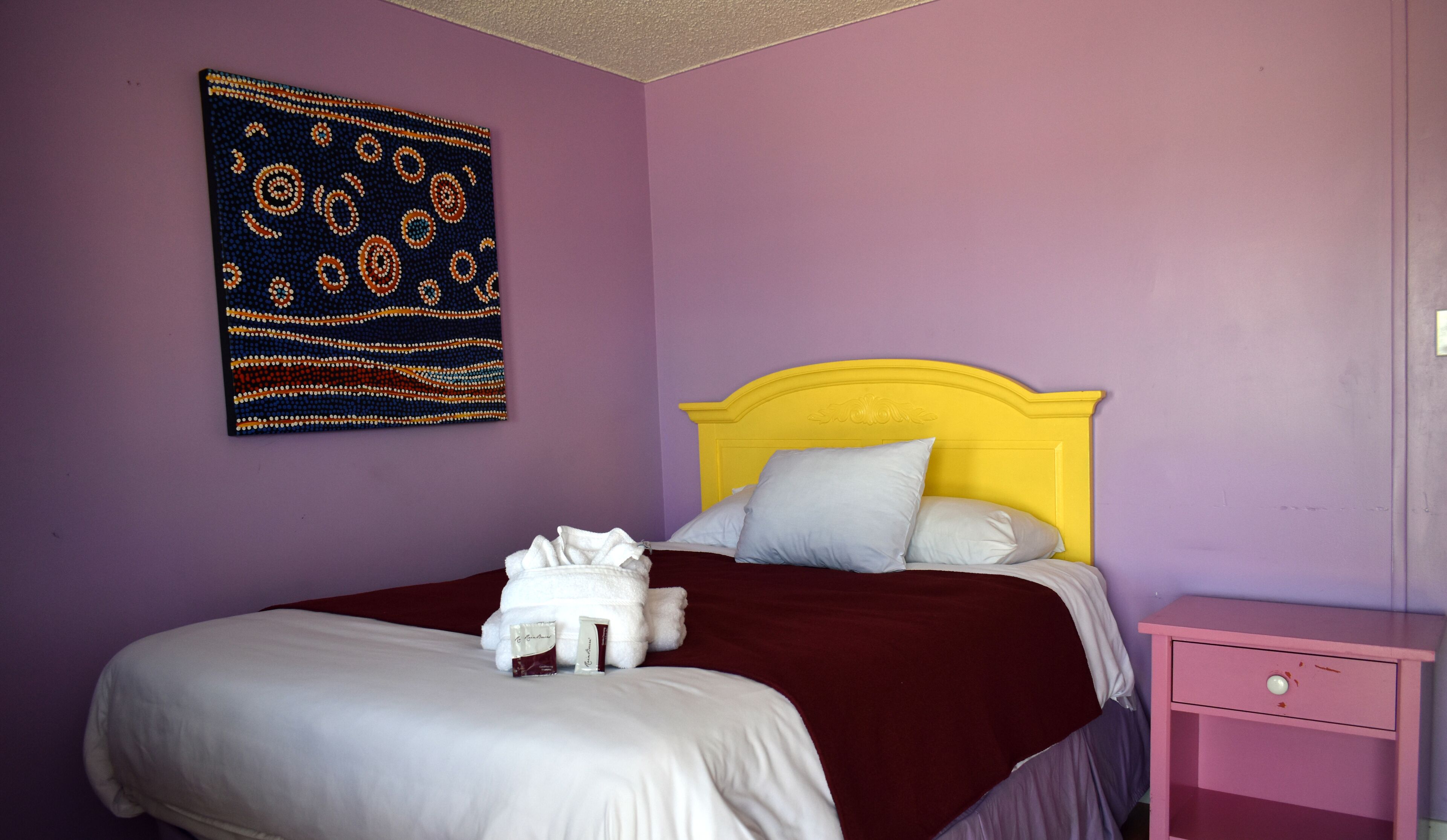 Cama com cabeceira amarela, roupa de cama branca e um cobertor decorativo.