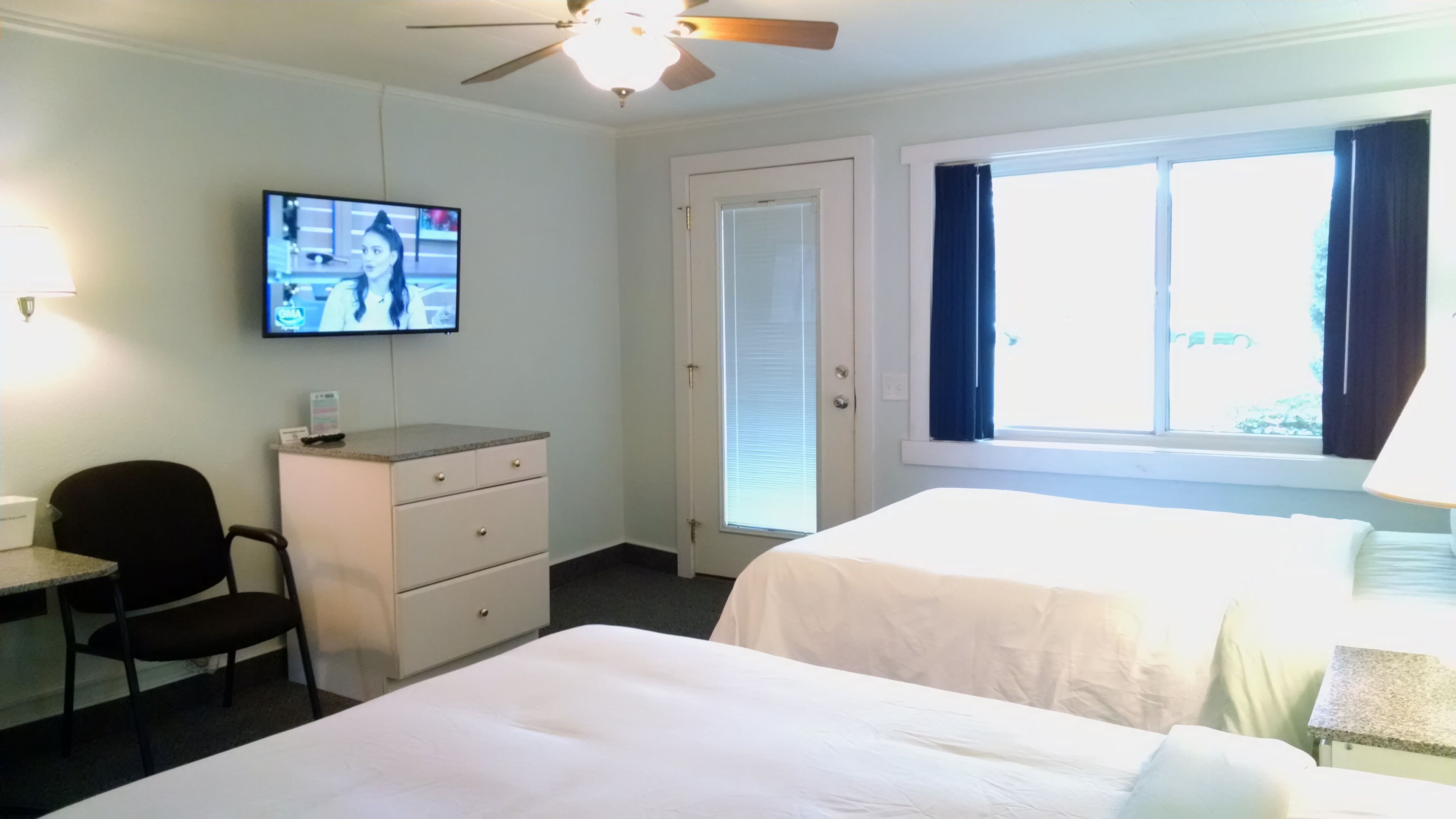 Quarto de hotel com cama, escrivaninha, cadeira, televisão e janela com cortinas.