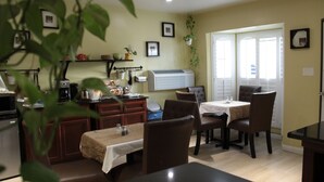 Uma sala de jantar com uma mesa posta para quatro pessoas, rodeada por cadeiras. Dá para ver uma cozinha ao fundo, com fogão e armários.