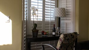 Quarto com janela com persianas, planta em vaso, tigela de frutas, garrafa de vinho, copo e cadeira estampada.