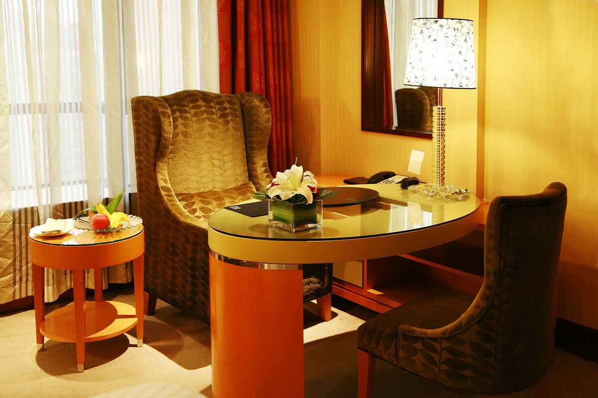 Quarto de hotel com uma mesa de vidro redonda, duas cadeiras, uma mesinha de apoio com abajur e uma cama com cortinas listradas.
