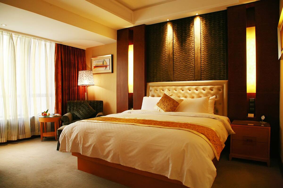 Quarto de hotel com cama grande, uma cadeira, mesa de cabeceira e abajur.