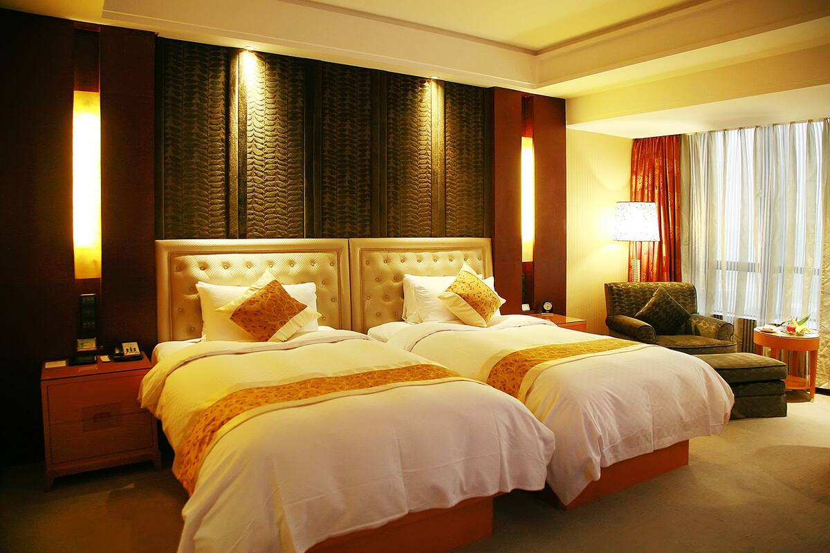Quarto de hotel com duas camas grandes, uma mesinha de cabeceira, uma cadeira e uma pequena mesa.